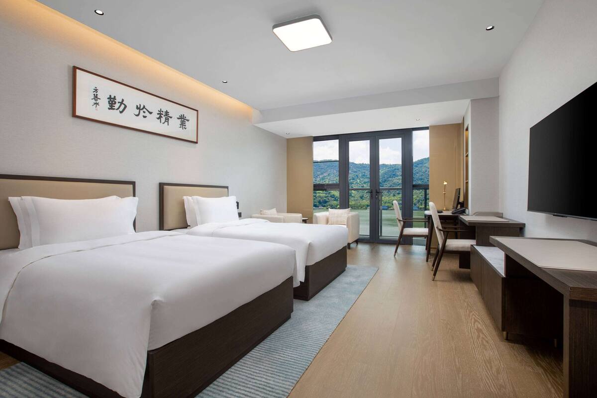 Quarto de hotel com duas camas, uma escrivaninha, uma cadeira e uma televisão. Há uma janela grande com vista para o exterior.