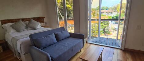 Quarto com cama, sofá azul, mesa de madeira e vista para uma varanda com mobiliário externo.