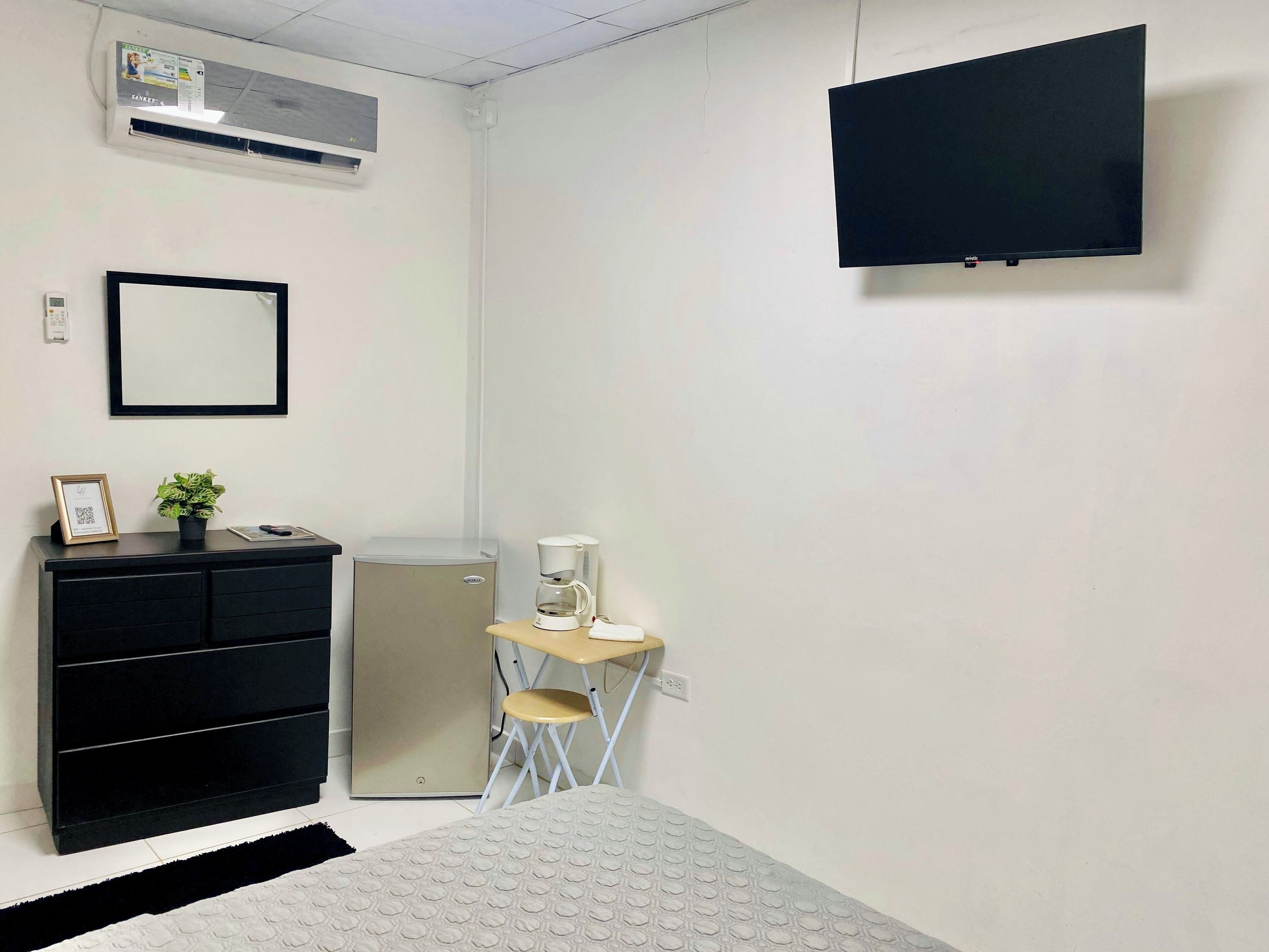 Quarto com guarda-roupa preto, uma mesa pequena com máquina de café, planta em vaso, quadro emoldurado, espelho e televisão na parede.