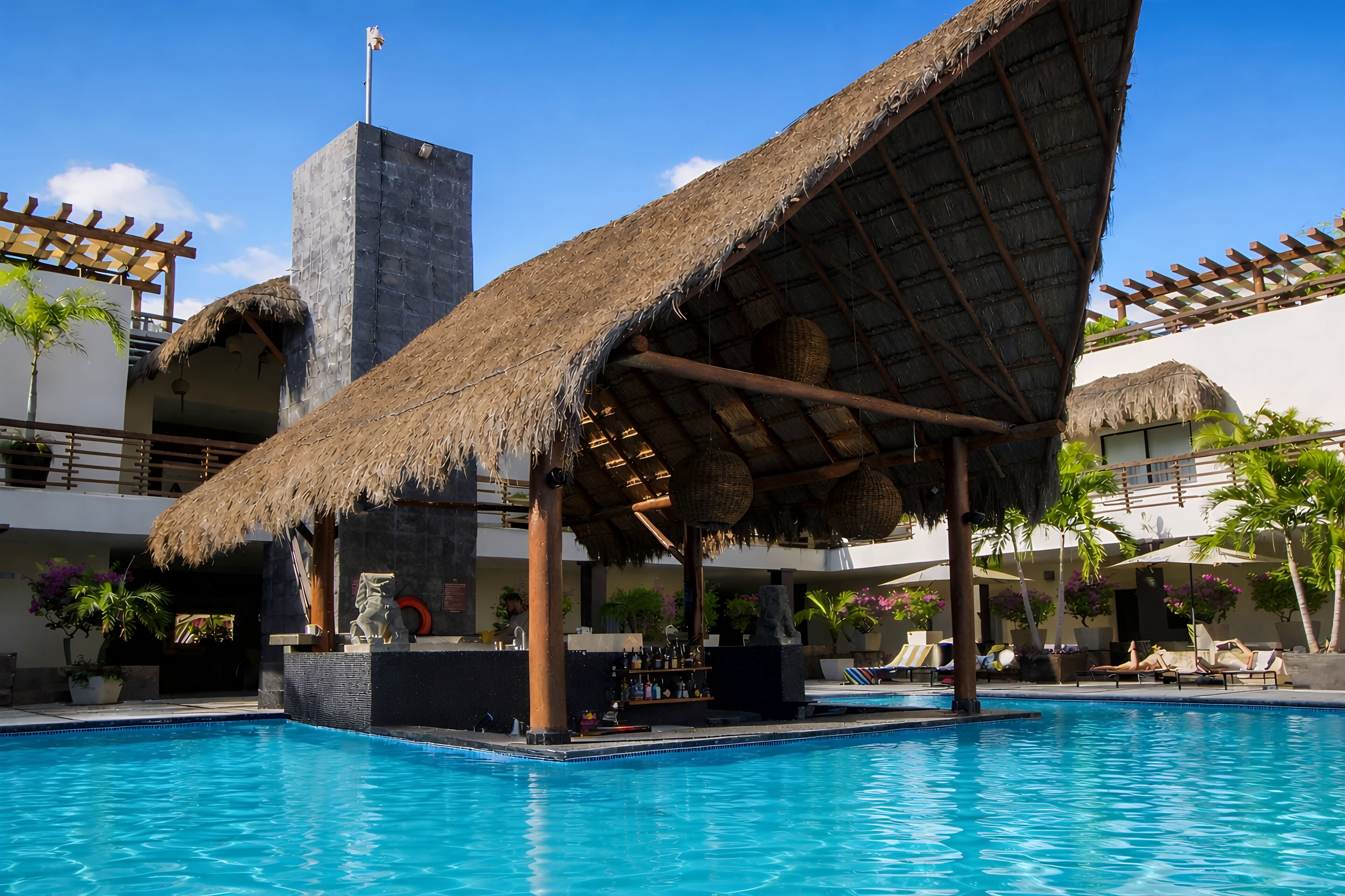 Uma área de piscina com estrutura de telhado de palha, espreguiçadeiras e um bar.