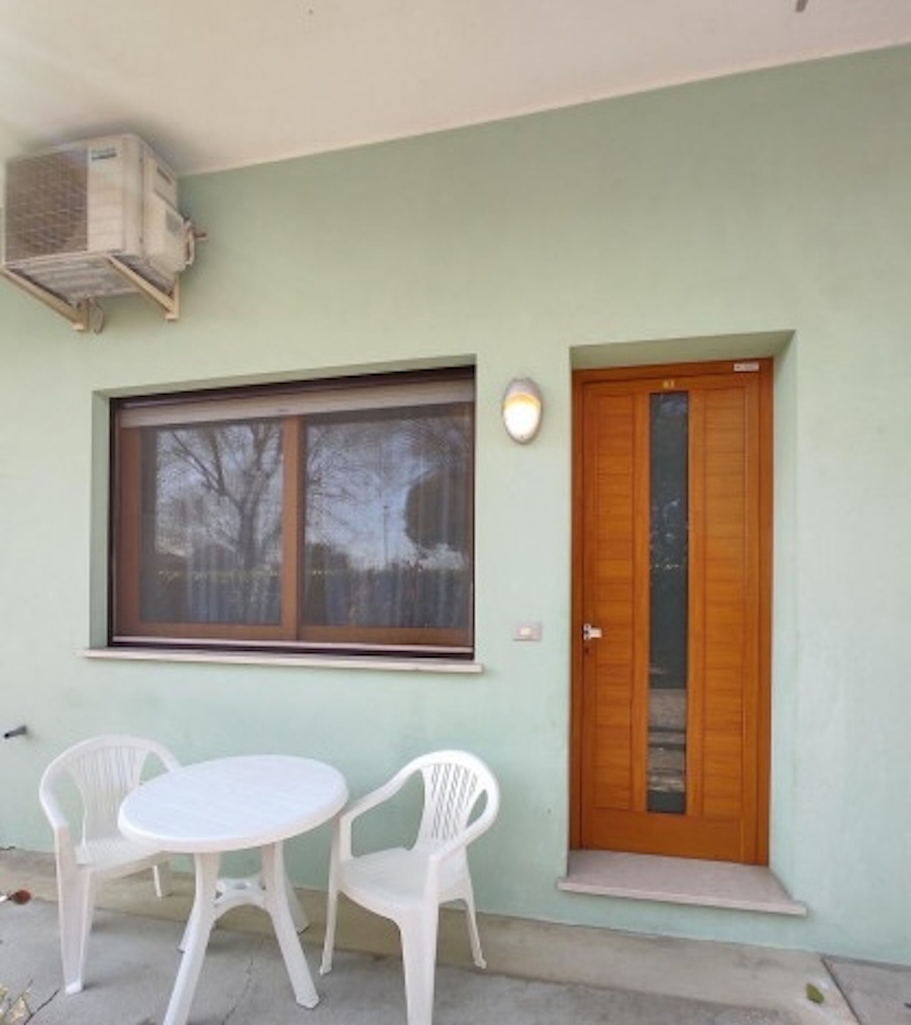 Um edifício com uma janela grande, uma porta e um pequeno alpendre com duas cadeiras de plástico branco.