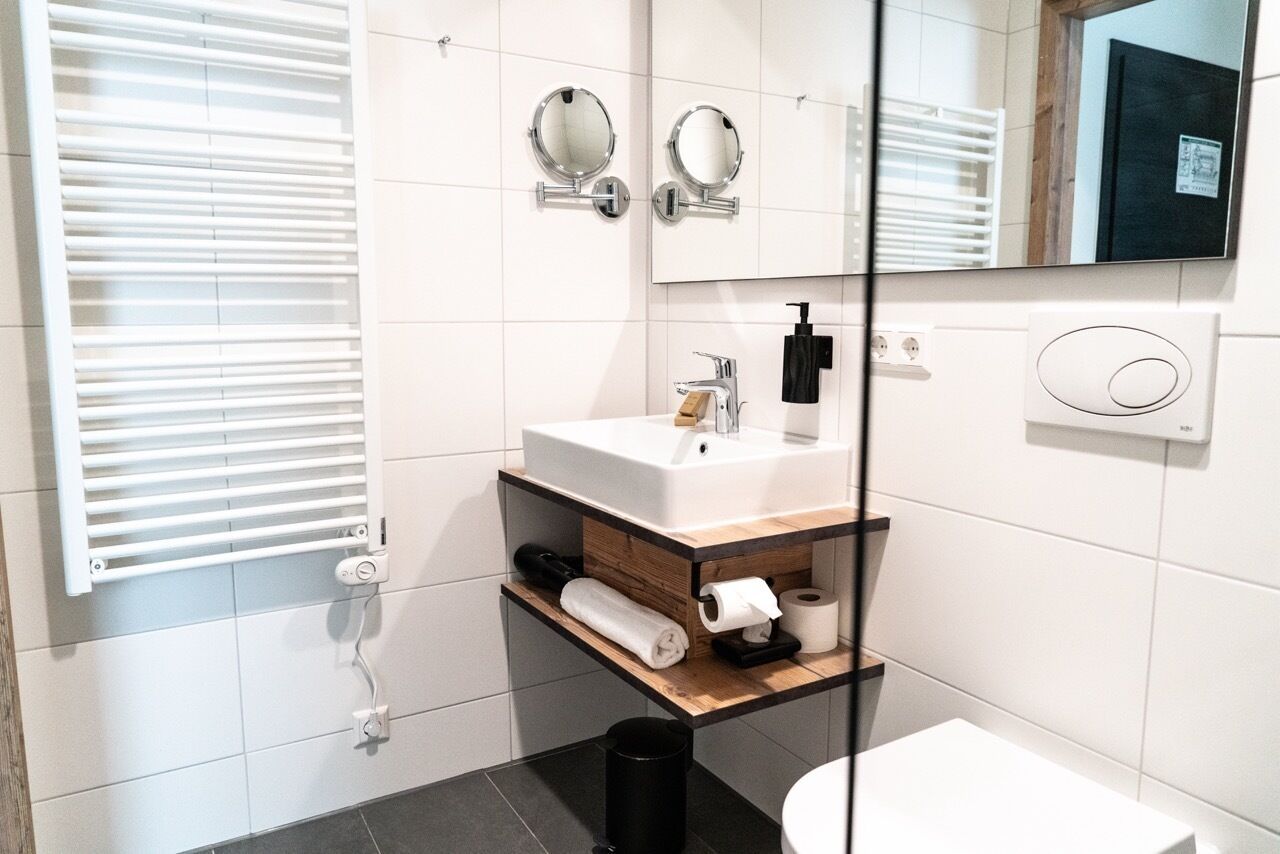 Uma casa de banho moderna com uma pia branca, dois espelhos redondos, uma prateleira de madeira com artigos de higiene e um cesto de lixo preto, e uma sanita.