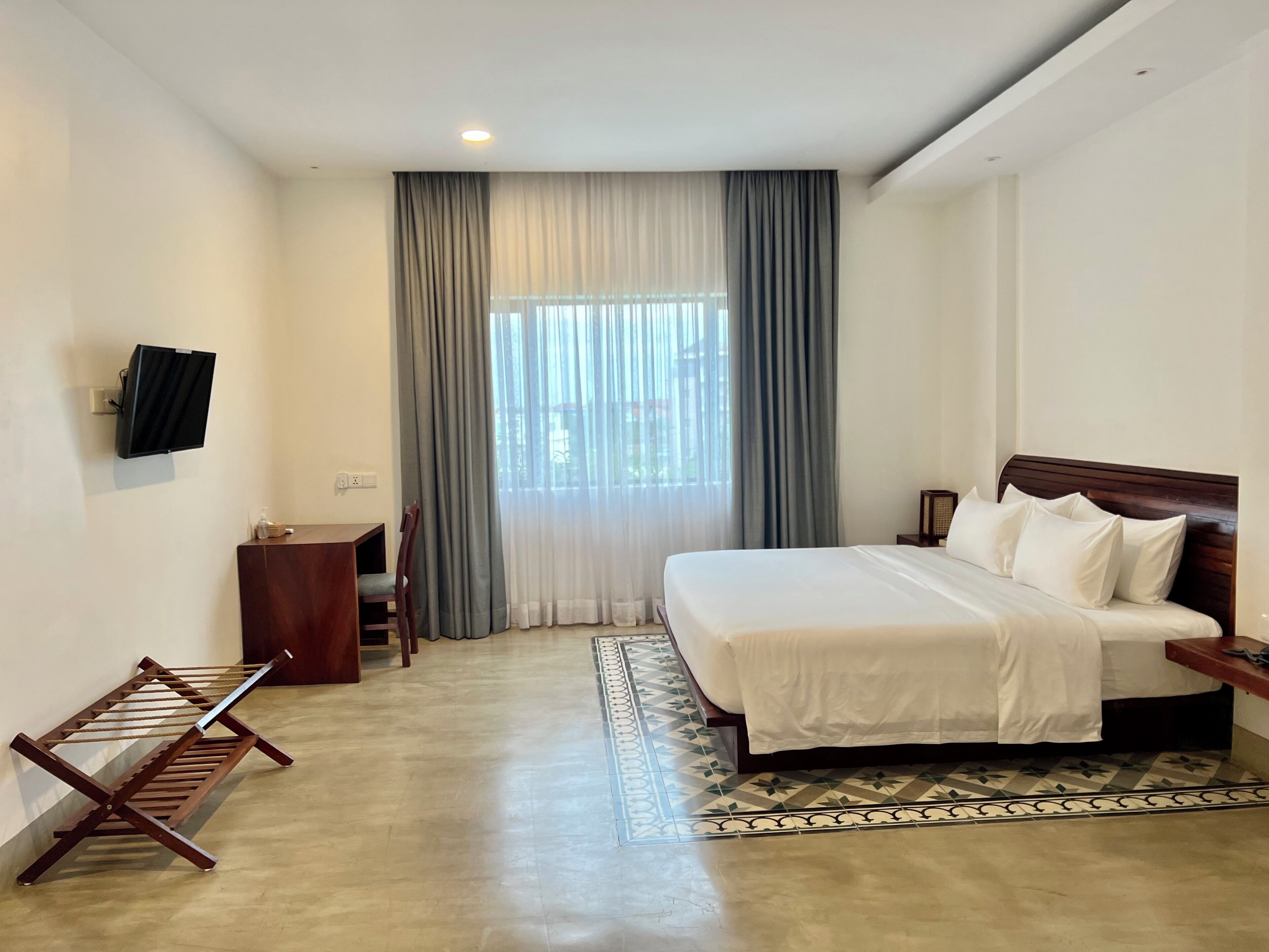 Quarto de hotel com uma cama, televisão na parede, escrivaninha com cadeira e janela com cortinas.