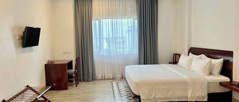 Quarto de hotel com uma cama, televisão na parede, escrivaninha com cadeira e janela com cortinas.