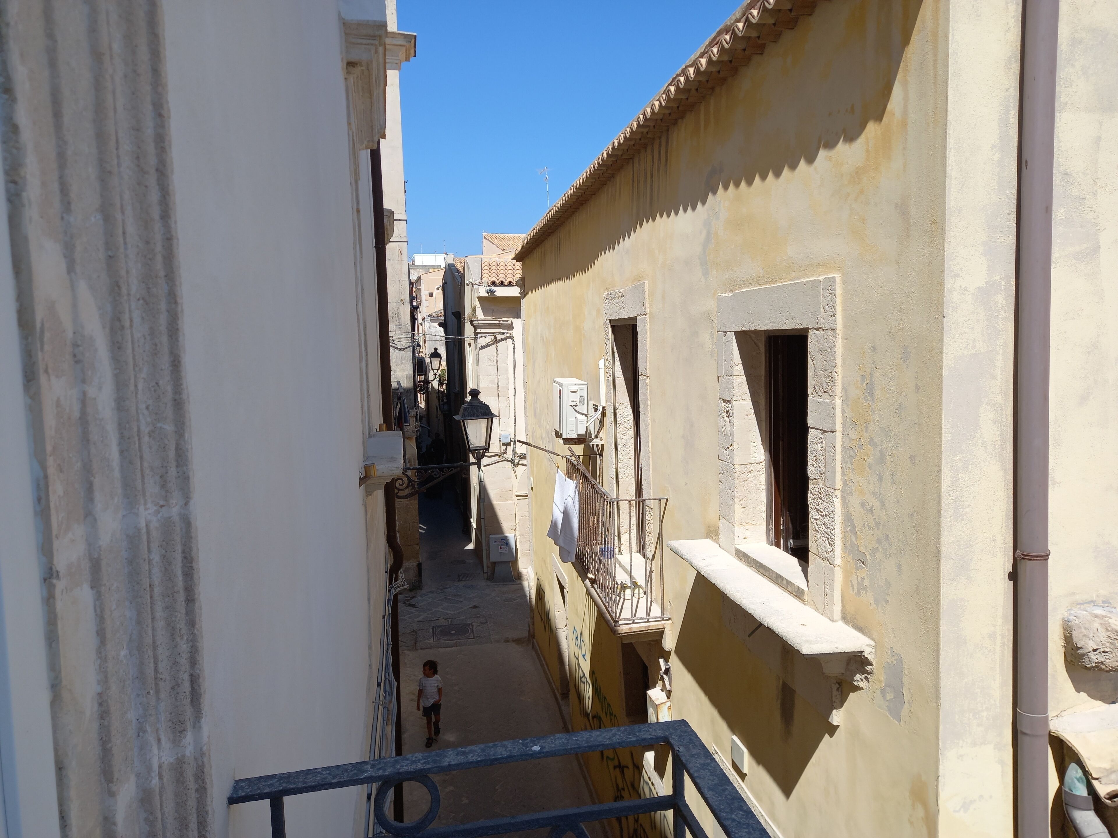 Uma viela estreita com edifícios em ambos os lados, uma criança caminhando pela passagem e um corrimão azul em primeiro plano.