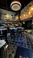 Uma área de bar com diversas opções de assento, incluindo mesas e cadeiras, e um painel decorativo com padrão.