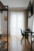 Quarto de hotel moderno com uma cama, uma escrivaninha com cadeira, televisão de tela plana na parede e uma janela grande com cortinas leves.