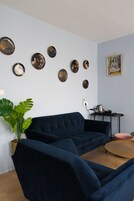 Sala de estar com um sofá azul escuro, uma mesa de centro redonda e uma planta. Há pratos decorativos pendurados na parede e um quadro.