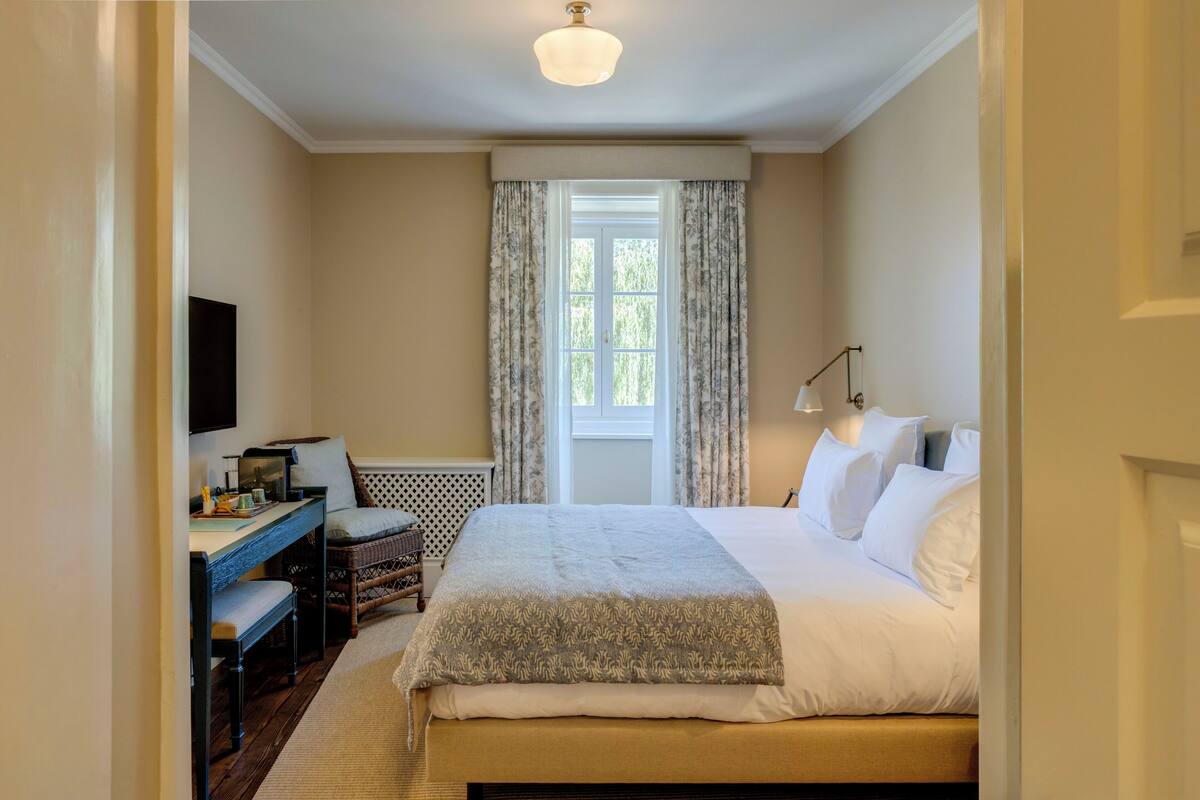 Quarto com cama, escrivaninha, cadeira, televisão e janela com cortinas.