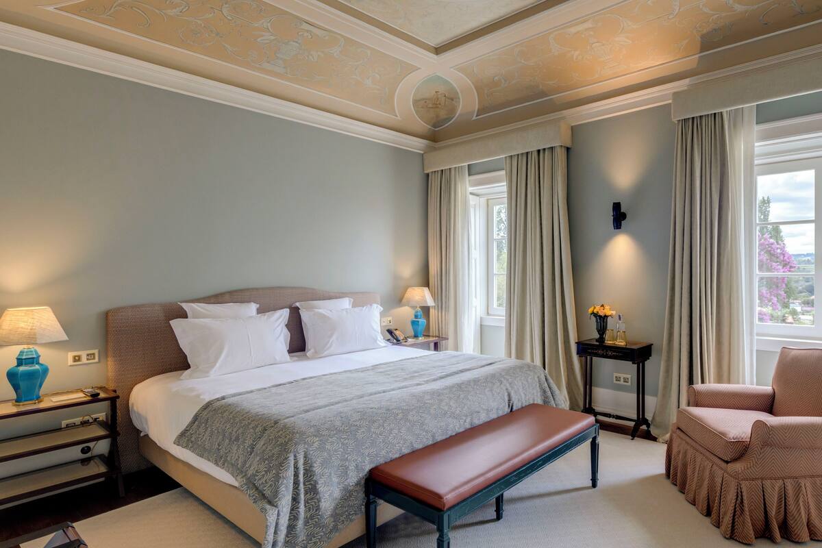 Quarto de hotel bem equipado com uma cama grande, um banco, uma cadeira, uma mesinha de cabeceira com abajures e vista para o exterior.