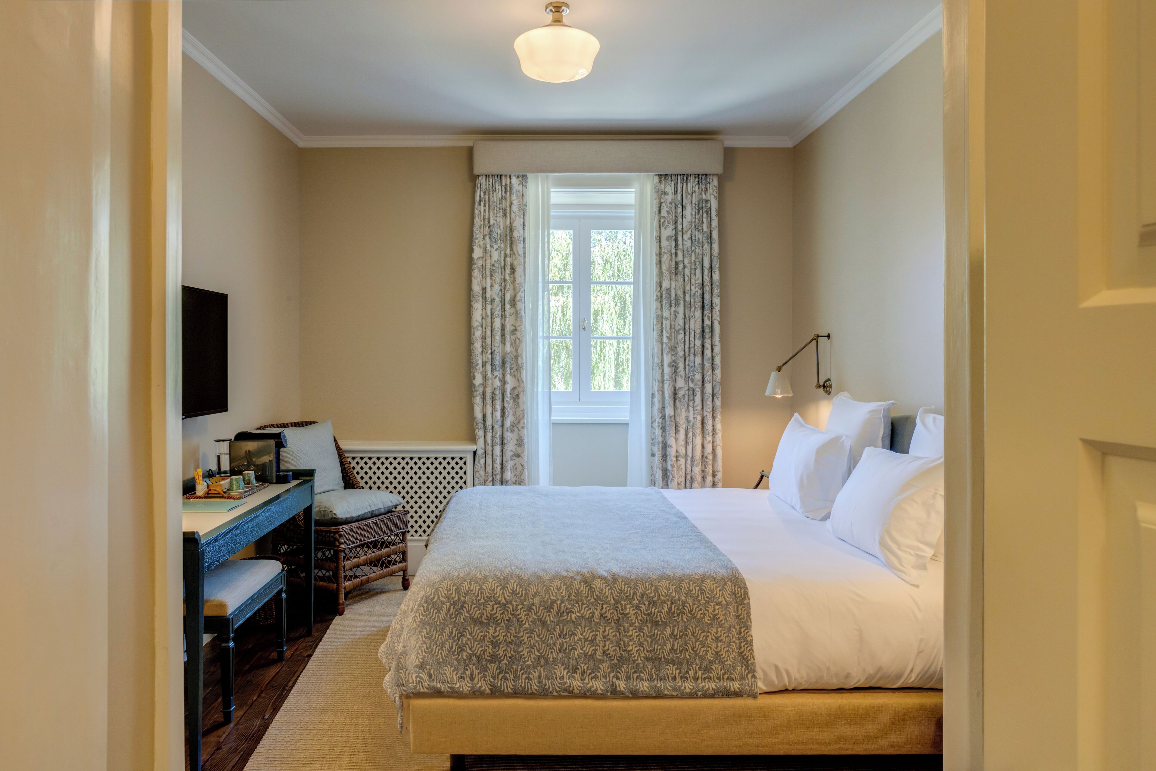 Quarto com cama, escrivaninha, cadeira, televisão e janela com cortinas.