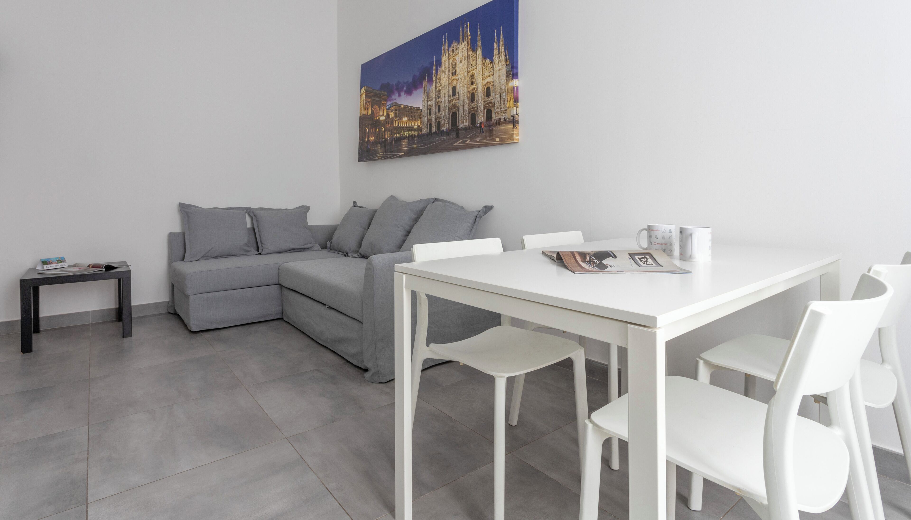 Uma sala moderna com um sofá modular cinza, uma mesa de jantar branca com cadeiras, uma pequena mesa lateral e um quadro de uma catedral na parede.