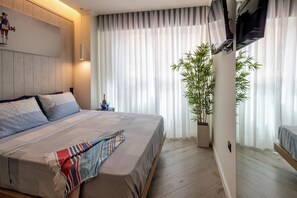 Quarto moderno com cama grande, mesa de cabeceira com abajur, planta em vaso e televisão na parede.