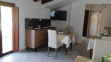 Uma área de refeições com mesa e cadeiras de madeira, bancada na cozinha com uma TV e uma porta que dá para outro quarto.