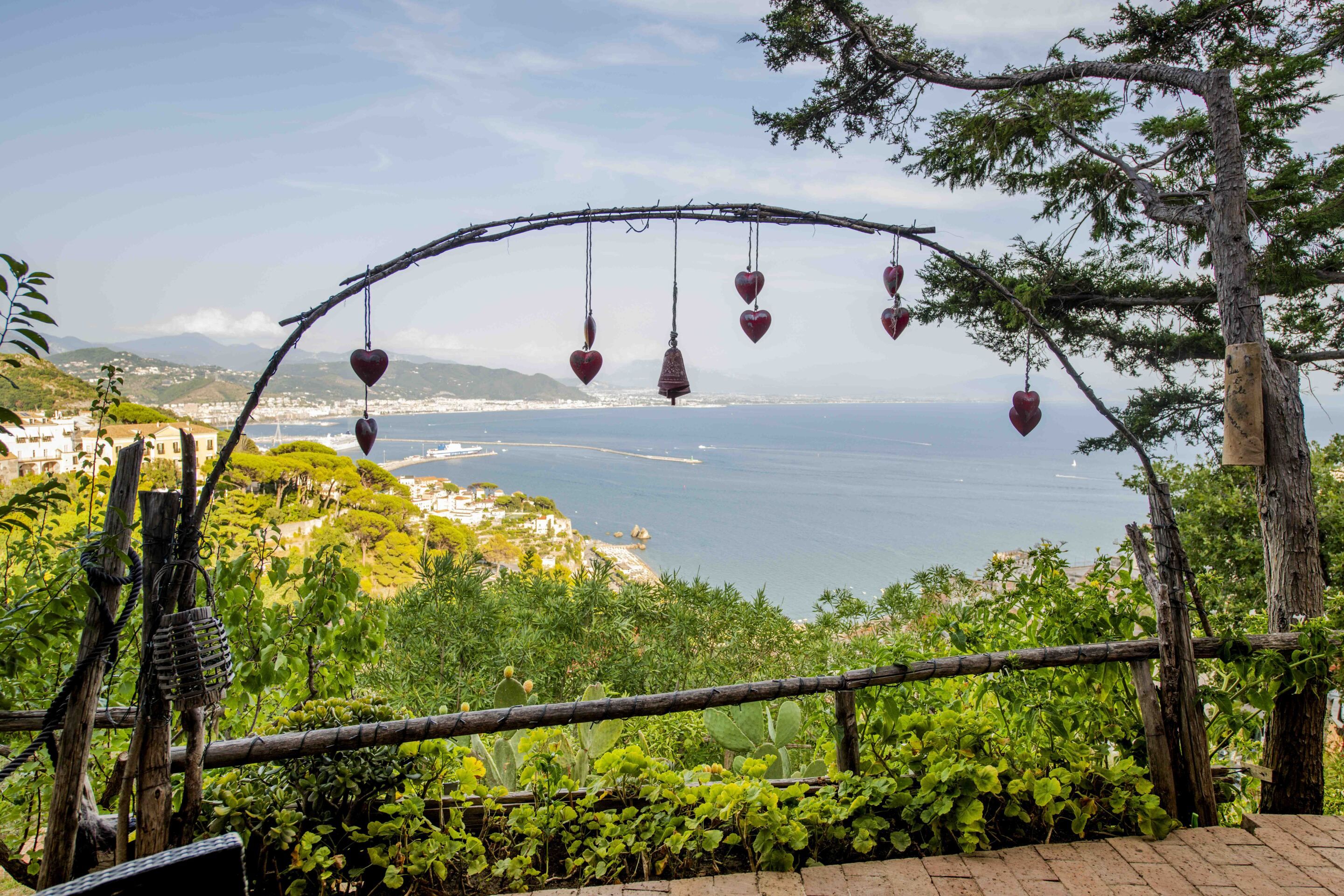 Vista de uma paisagem costeira com um arco de madeira decorado com corações, que dá acesso a uma área verde exuberante com cactos e uma visão clara do mar e colinas ao longe.