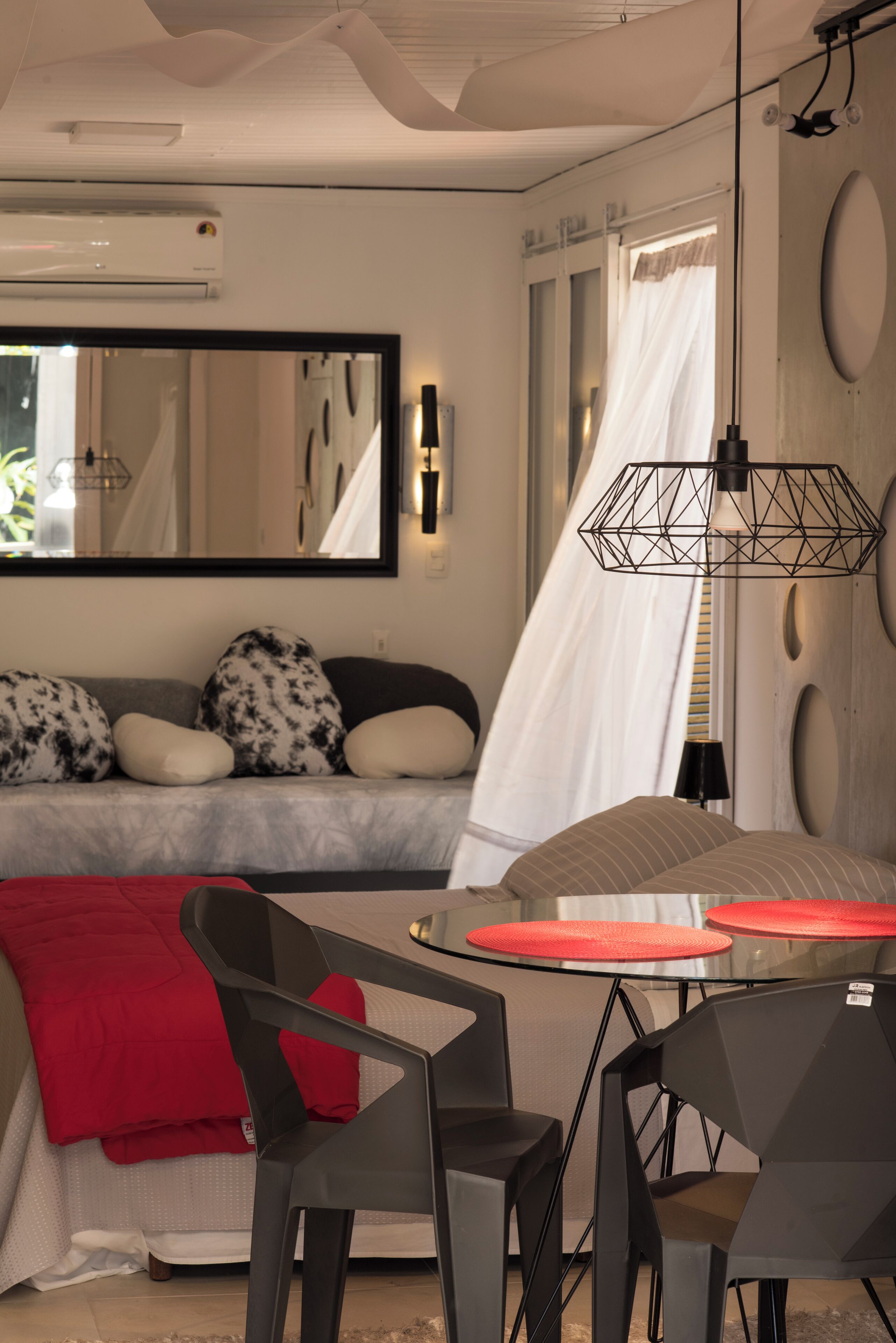 Quarto moderno com cama, espelho grande, luminária pendente e mesa de vidro com cadeiras.