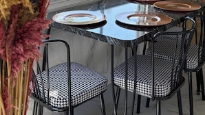 Uma área de refeições com uma mesa posta para quatro pessoas, rodeada por cadeiras, perto de uma janela com persianas.