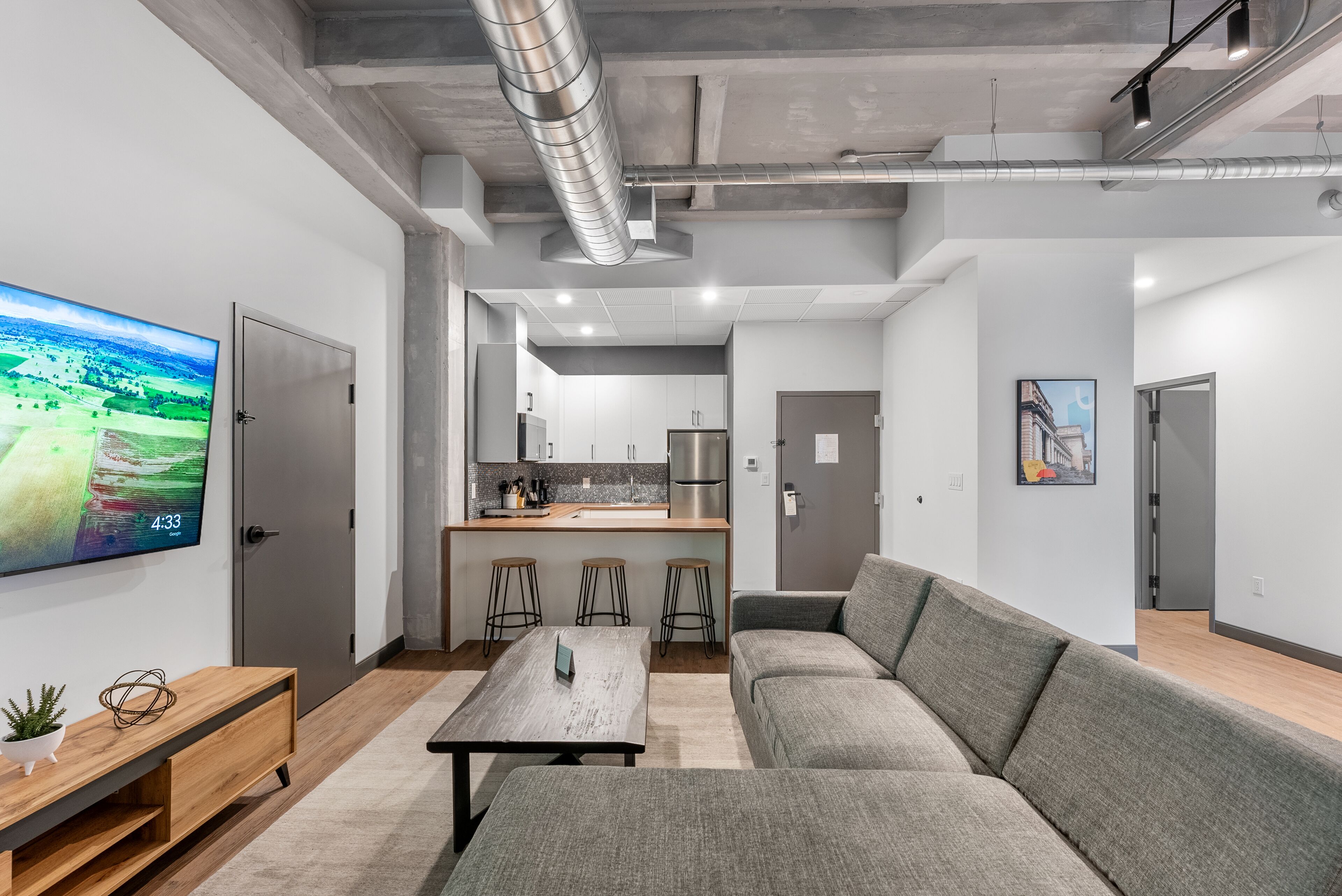 Uma sala de estar moderna com uma televisão de tela plana grande, um sofá cinza, uma mesa de centro de madeira e uma área de cozinha ao fundo.