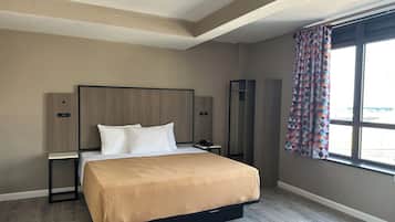 Quarto de hotel com cama, mesa de cabeceira, espelho, janela com cortinas e piso de madeira.