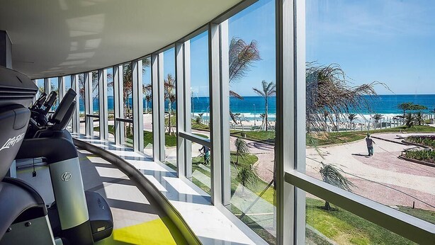 Academia com equipamento de exercícios e amplas janelas com vista para a praia.