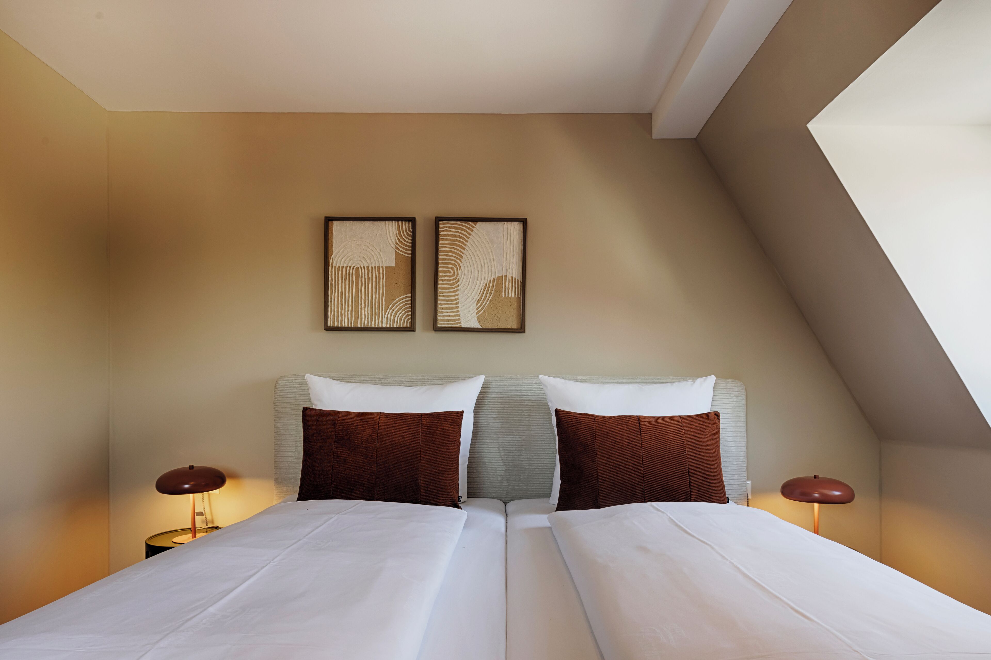 Quarto de hotel com duas camas, teto inclinado e duas obras de arte na parede.
