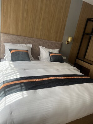 Uma cama bem arrumada com roupa de cama branca e cinza, cabeceira de madeira e uma luminária fixada na parede.
