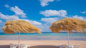 Dois guarda-sóis e duas cadeiras de praia brancas numa praia arenosa, com água azul cristalina e céu ensolarado.