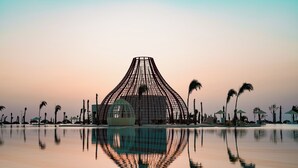 Um edifício moderno com um design geométrico único, cercado por palmeiras e uma piscina calma e espelhada.