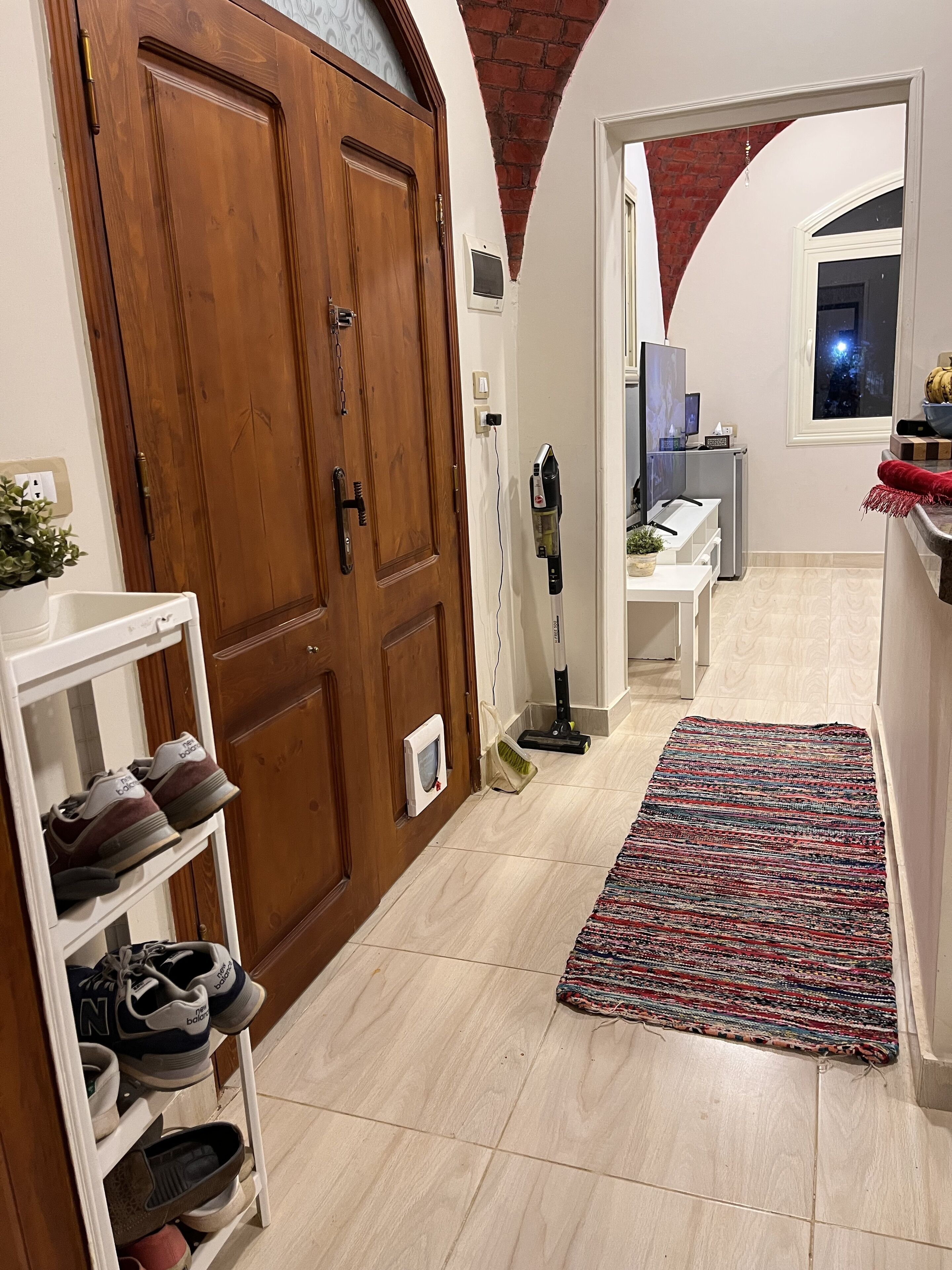 Um corredor com porta de madeira, sapateira, aspirador de pó, tapete estampado e janela com vista noturna.