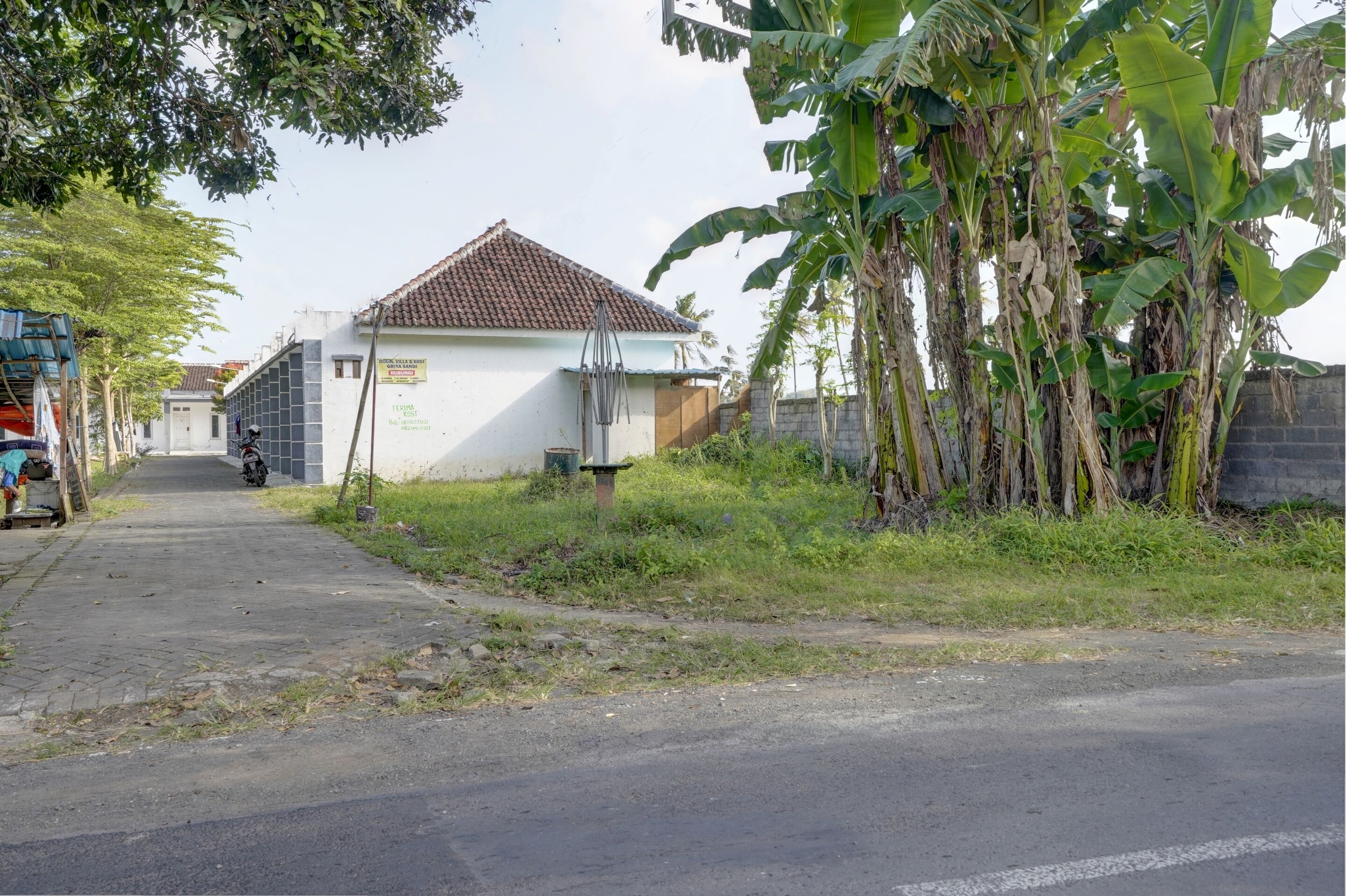 Um edifício residencial com telhado telhado, pequena área de jardim com bananeiras, e caminho pavimentado que leva à entrada.