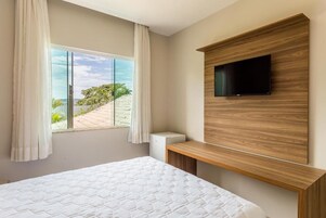 Quarto com uma janela grande, uma cama, uma TV fixada em um painel de madeira e um armário pequeno branco.