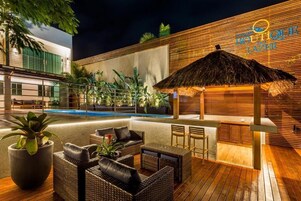 Uma área de piscina moderna ao ar livre, com mobiliário de lounge, um bar e um guarda-sol de palha.
