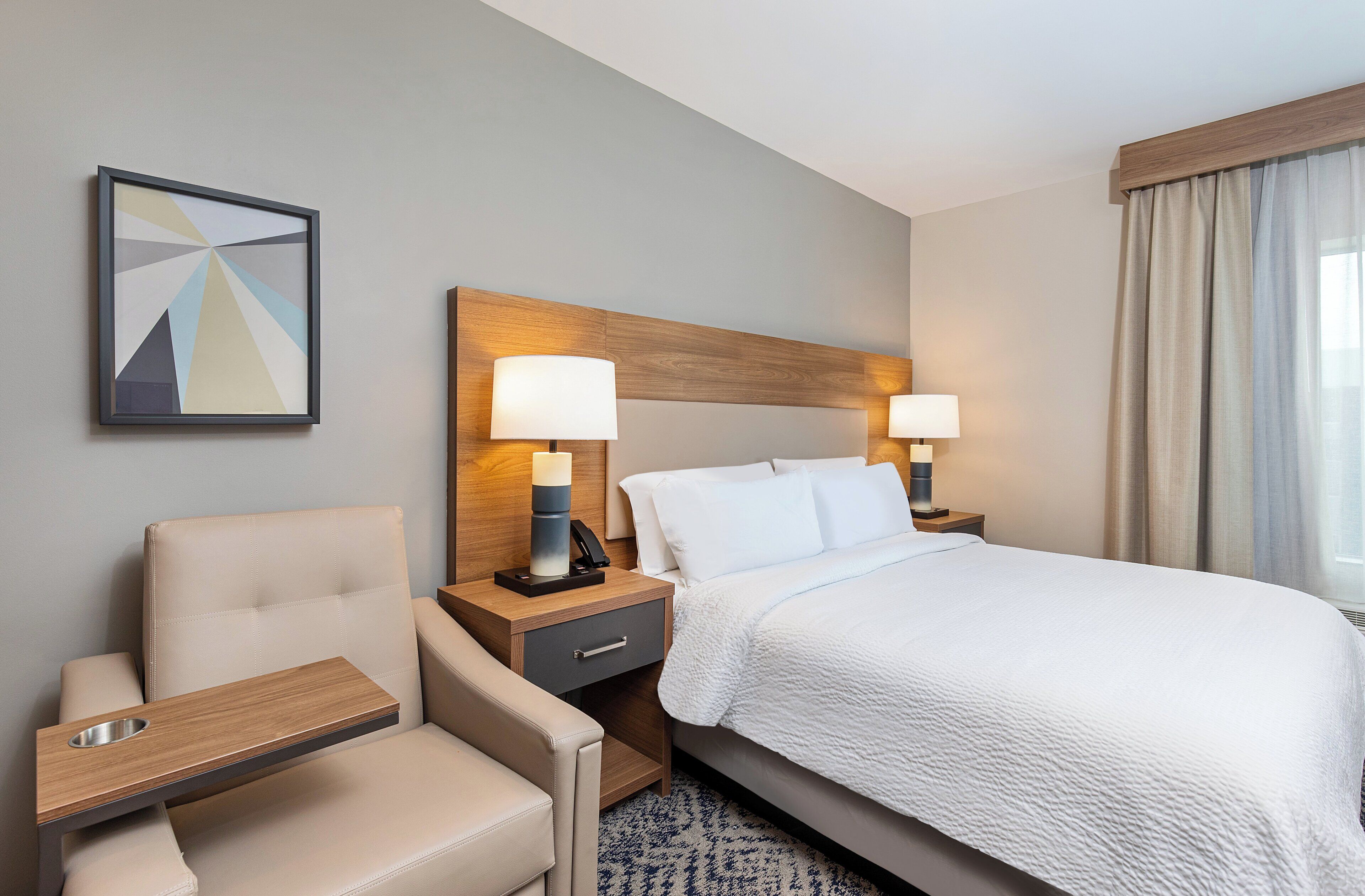 Quarto de hotel com cama, duas mesas de cabeceira com abajures, uma cadeira, uma mesinha e uma obra de arte na parede, além de uma janela com cortinas.