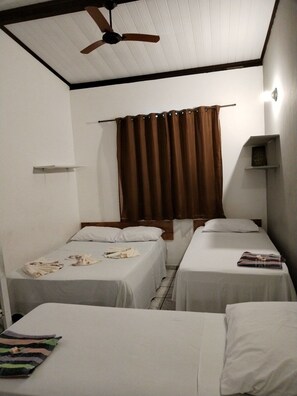 Um quarto com duas camas de solteiro, um ventilador de teto, uma janela com cortinas castanhas e uma pequena prateleira com um livro.
