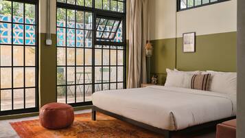 Quarto com cama grande, janela com vista para vegetação, tapete estampado e pequena mesa com abajur.