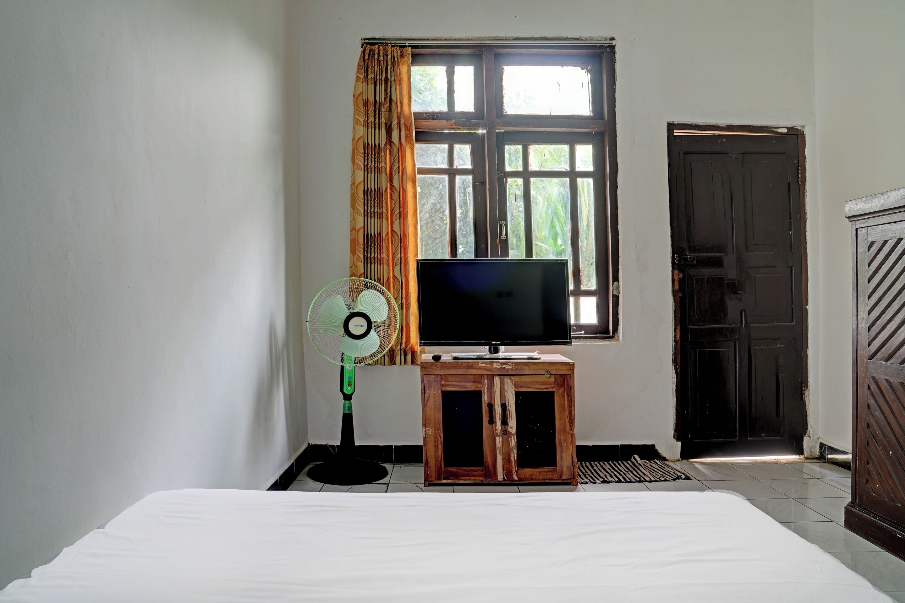 Quarto com cama, televisão sobre um móvel de madeira, um ventilador e uma janela com cortinas.