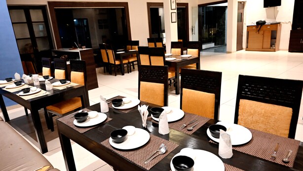 Uma área de refeições com várias mesas preparadas para o serviço, cadeiras e uma cozinha ao fundo.