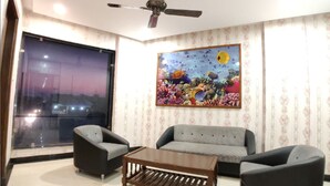 Uma sala de estar com ventilador de teto, dois poltronas, um sofá e uma mesa de centro. Há uma janela grande com vista para o horizonte urbano ao entardecer.