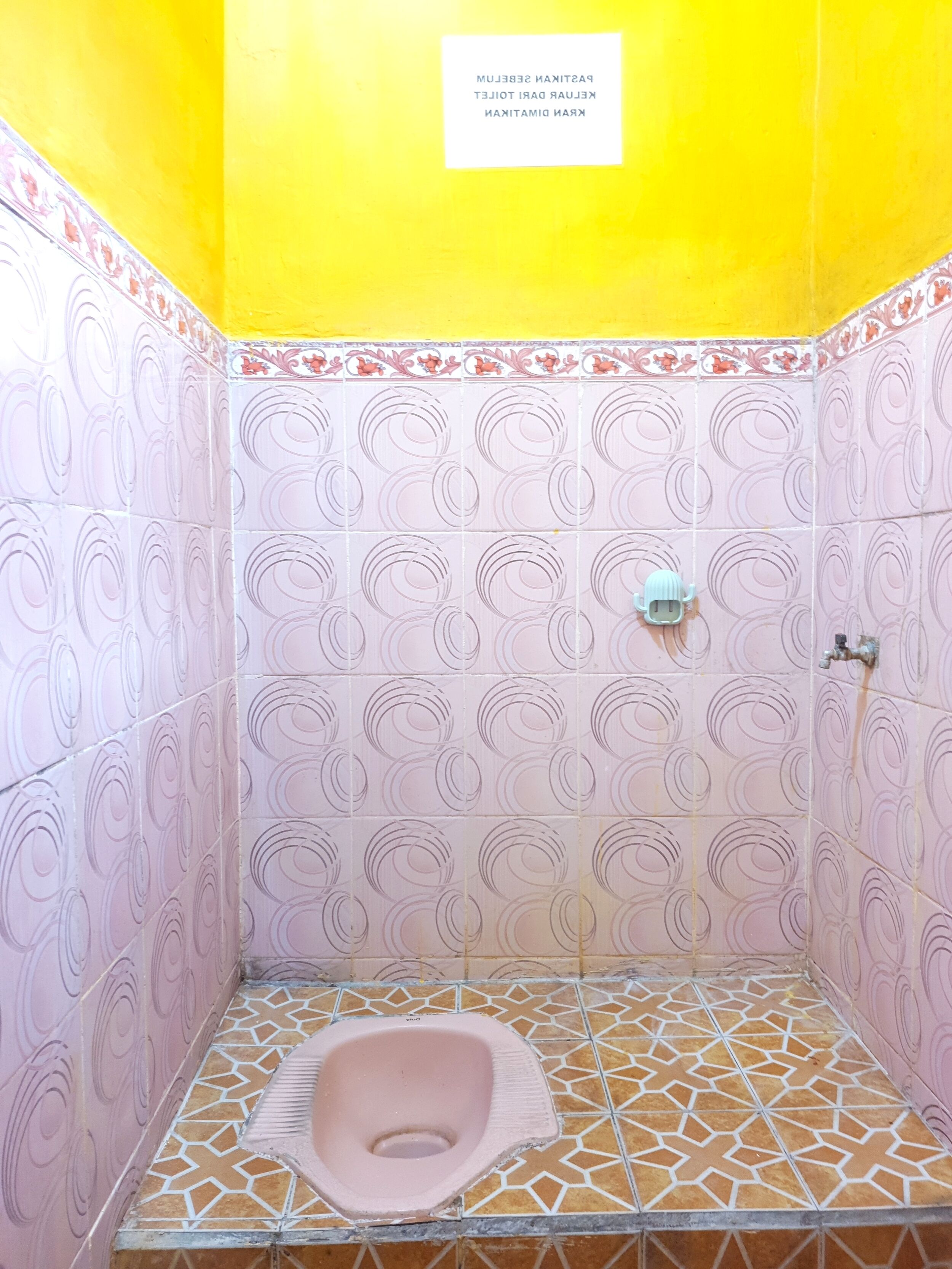Um banheiro pequeno com vaso sanitário, pia e chuveiro. As paredes são revestidas de azulejos com um padrão circular, e há uma placa sobre o vaso sanitário.
