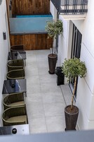 Um pátio exterior estreito com uma fileira de cadeiras e mesas de vime verde, duas plantas em vasos e uma piscina azul ao fundo.
