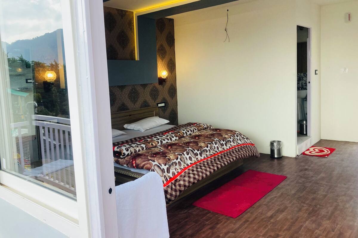 Quarto com cama, tapete vermelho, piso de madeira, parede estampada e varanda com guarda-corpo.