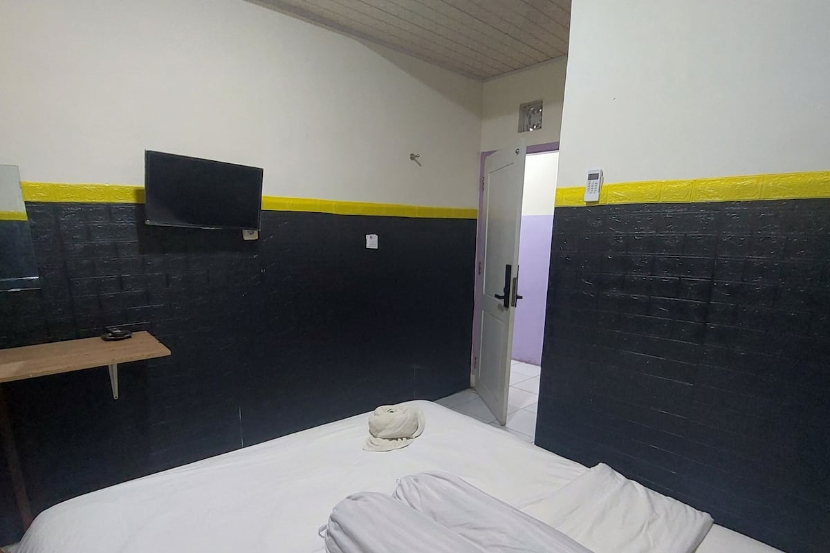 Quarto de hotel com cama, uma mesinha, televisão montada na parede e aparelho de ar condicionado.