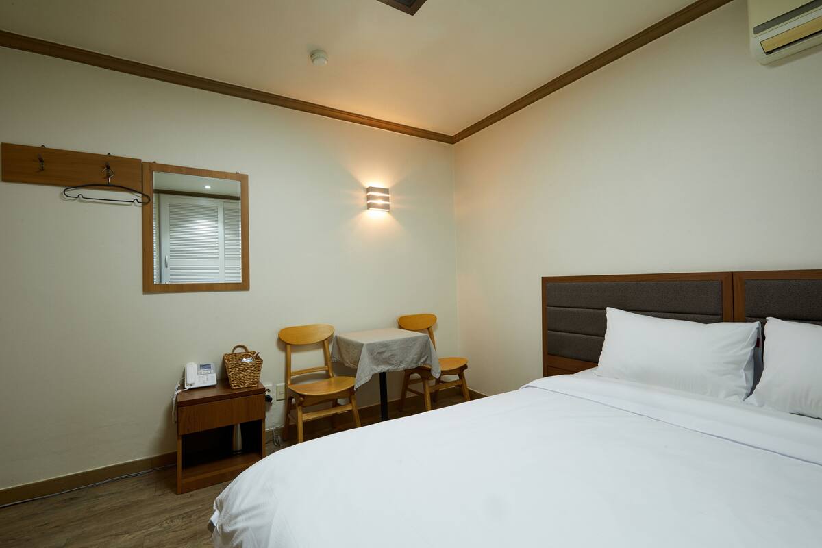 Quarto de hotel com cama, mesa de cabeceira, luminária, cadeira, mesinha e um cabideiro.