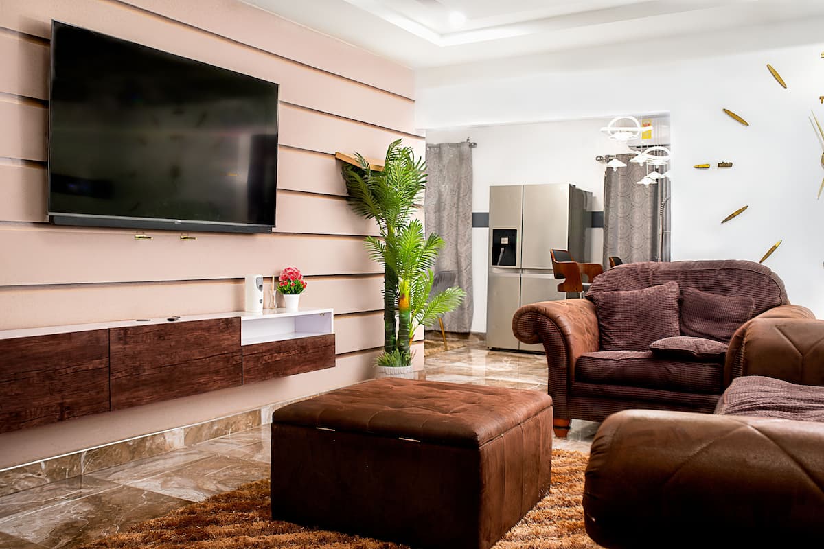 Uma sala de estar moderna com uma TV de tela plana na parede, um sofá de couro marrom, um banquinho combinando e um vaso de planta.