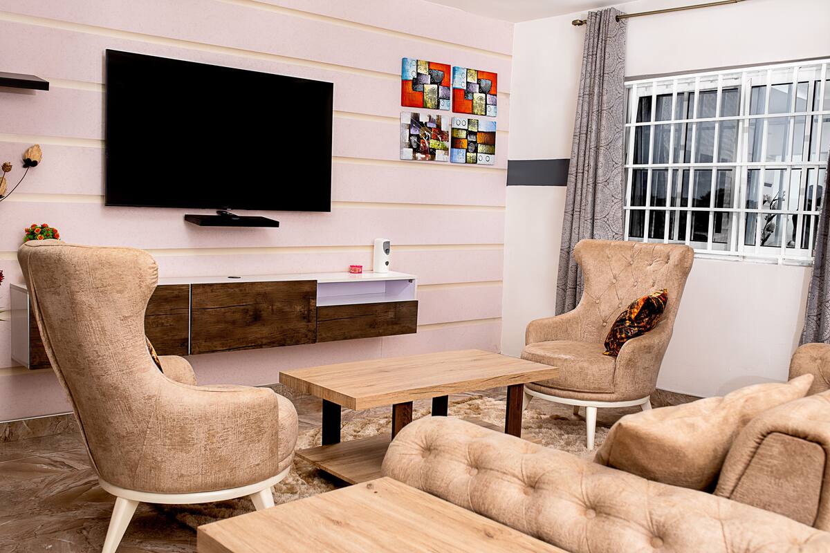 Uma sala de estar com televisão de tela plana, dois poltronas, um sofá, uma mesa de centro de madeira e uma estante fixada na parede.