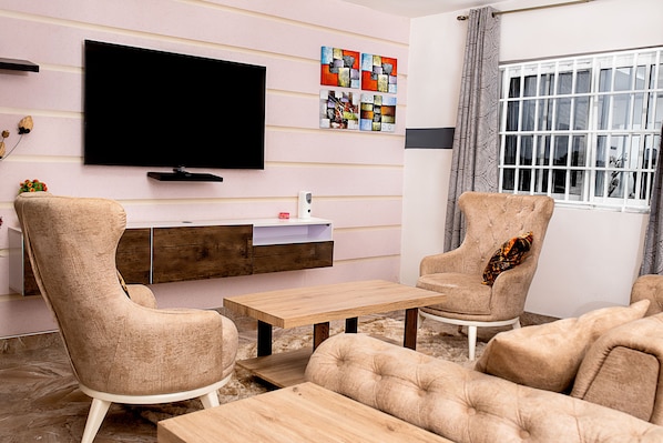 Uma sala de estar com televisão de tela plana, dois poltronas, um sofá, uma mesa de centro de madeira e uma estante fixada na parede.