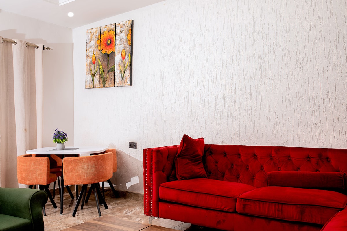 Uma sala de estar moderna com um sofá vermelho, poltronas laranjas, uma mesa branca e um quadro de flores na parede.