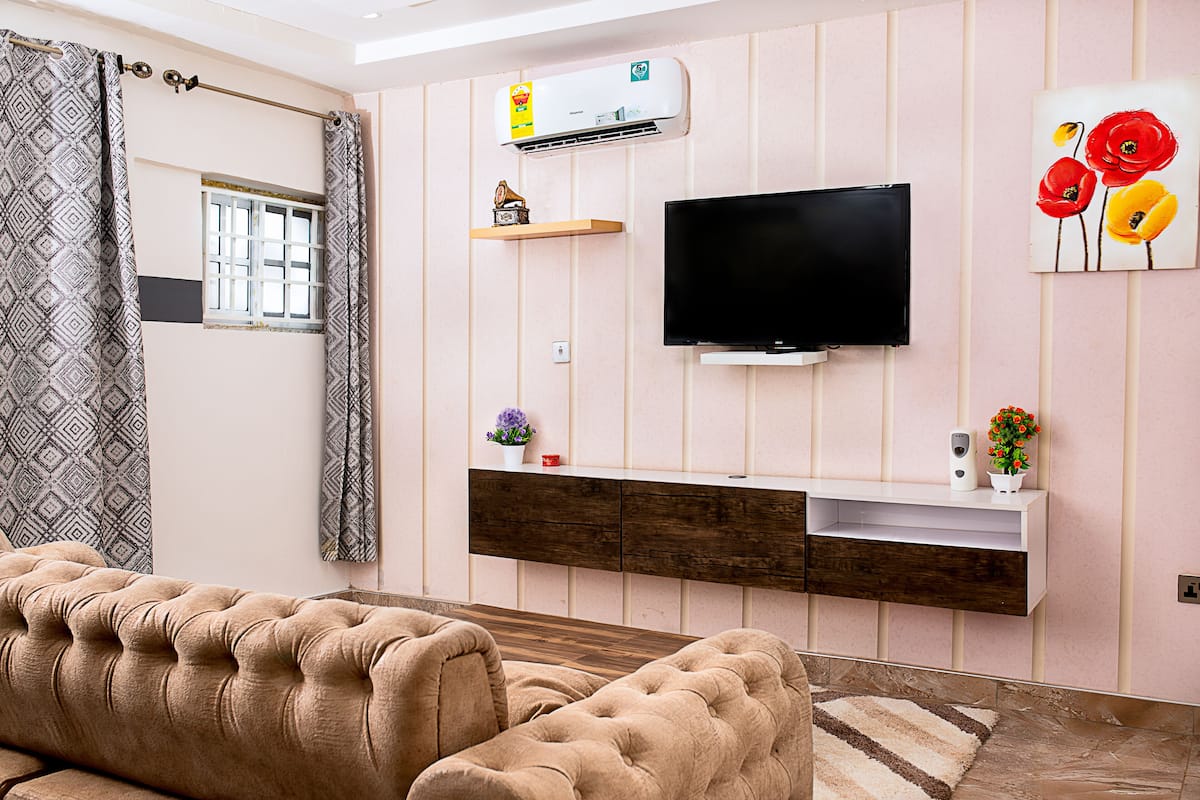 Uma sala de estar moderna com uma grande TV de tela plana fixada em um painel de madeira, um sofá estofado, uma cortina estampada e uma pequena janela com vista para o exterior.