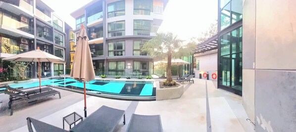 Área de piscina moderna ao ar livre, com espreguiçadeiras e guarda-sóis, cercada por edifícios de vidro.