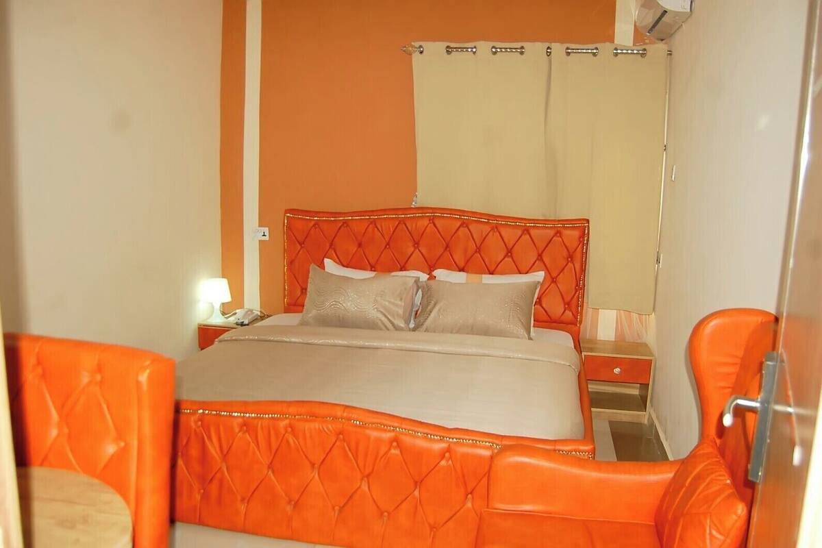 Quarto com uma cama, mesa de cabeceira, cadeira laranja e cortina.